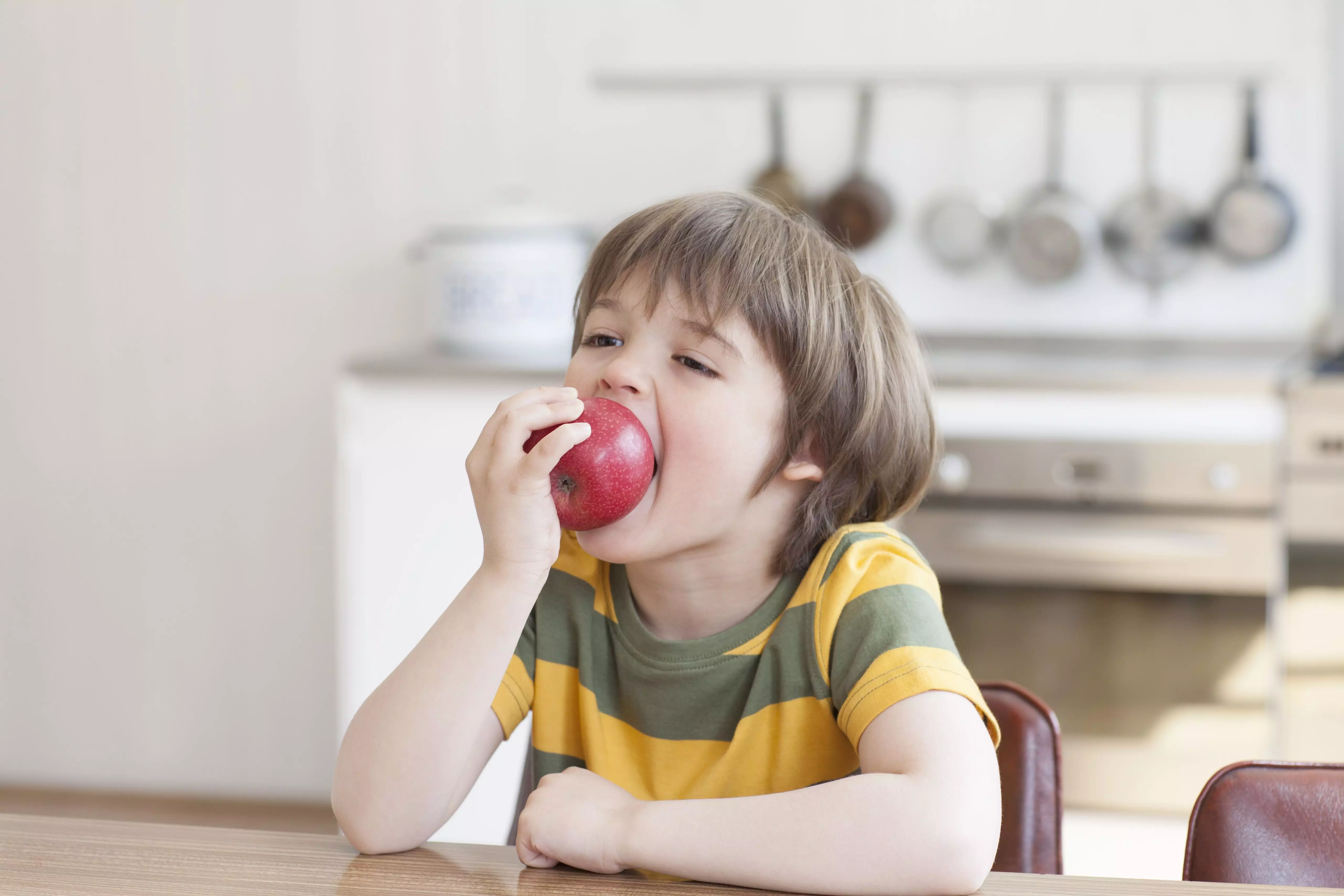 en liten pojke, som sitter vid ett bord, biter i ett rött äpple