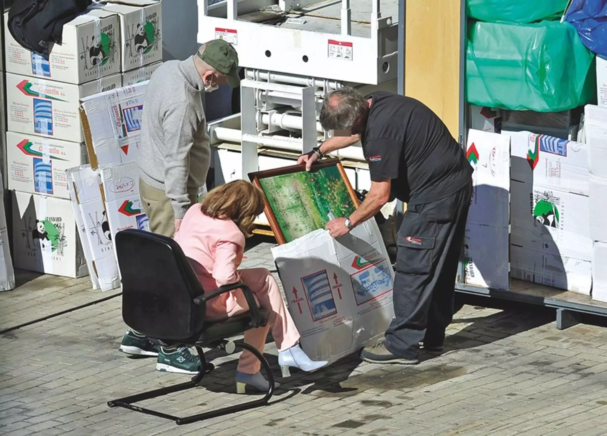 η παρουσιάστρια, καθισμένη σε μια πολυθρόνα, παρατηρεί έναν από τους πίνακές της με τη βοήθεια των εργαζομένων στην αποθήκευση επίπλων